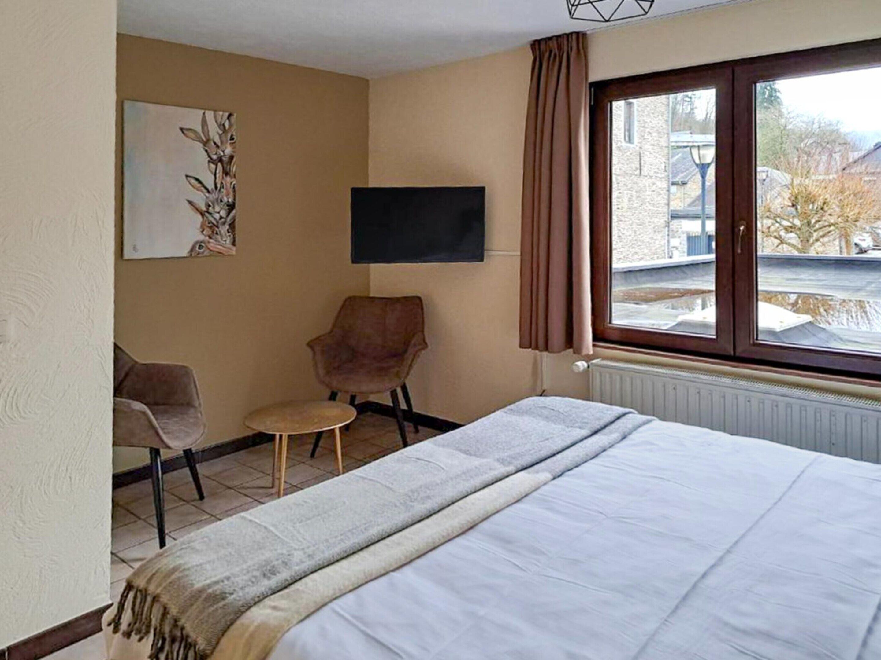 Quarto de hotel com uma cama, uma cadeira, uma mesinha, uma televisão e uma janela com cortinas.