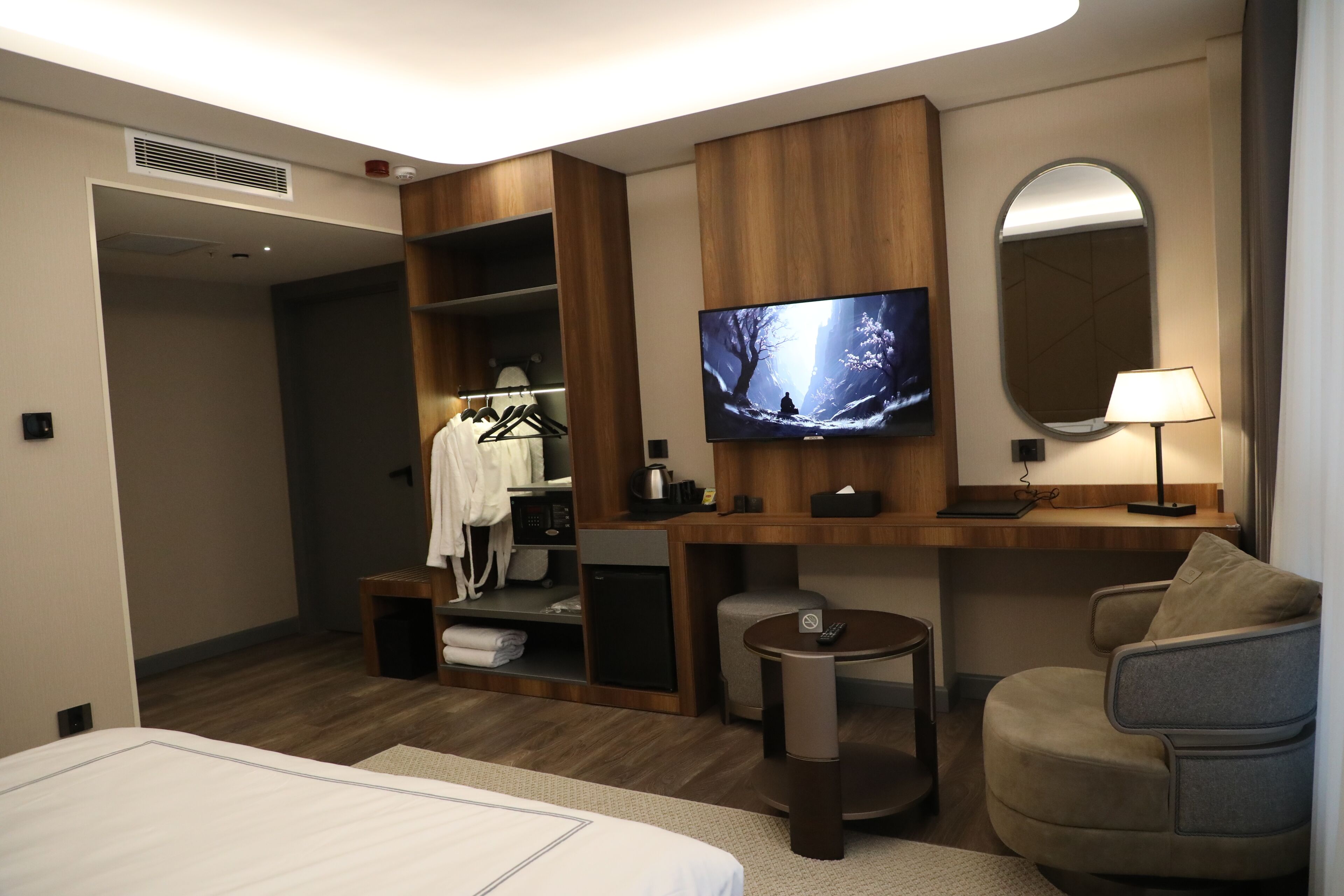 Quarto de hotel com cama, televisão, escrivaninha com luminária, cadeira, guarda-roupa e espelho.