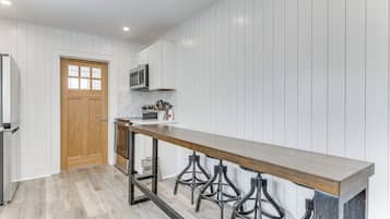 Uma cozinha com painéis de madeira branca, uma porta de madeira, uma geladeira de aço inoxidável, um micro-ondas, um balcão de madeira com banquetas e uma pia.