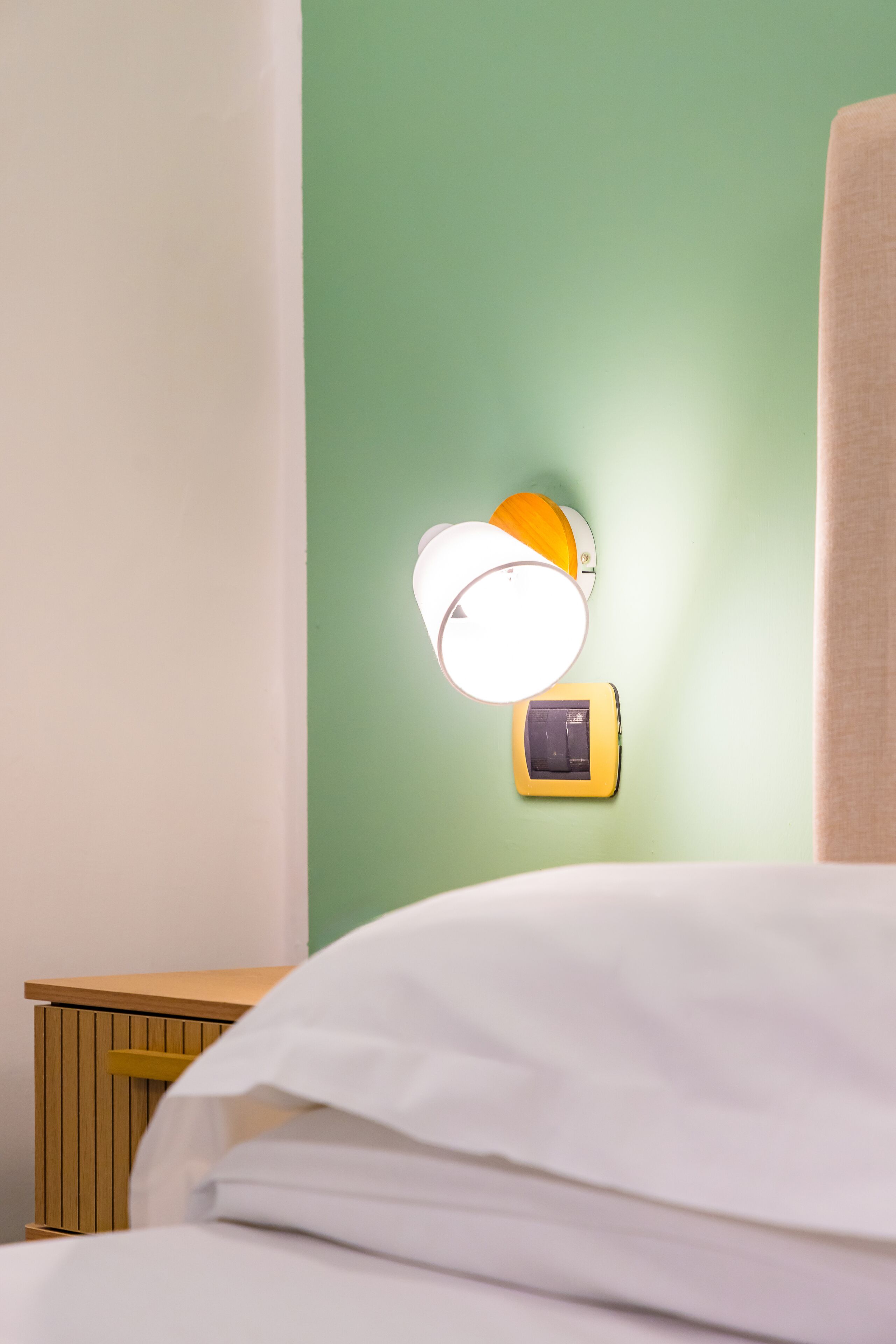 Cama bem arrumada com lençóis brancos, luminária fixada na parede e mesa de cabeceira de madeira.