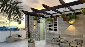 Terraço no telhado com pérgola, plantas em vasos, mesa com cadeiras e vista para a paisagem urbana ao pôr do sol.
