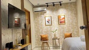 Quarto moderno com televisão de tela plana, ar condicionado, cama com roupa de cama branca, mesa pequena com planta e parede decorativa com obras de arte emolduradas.