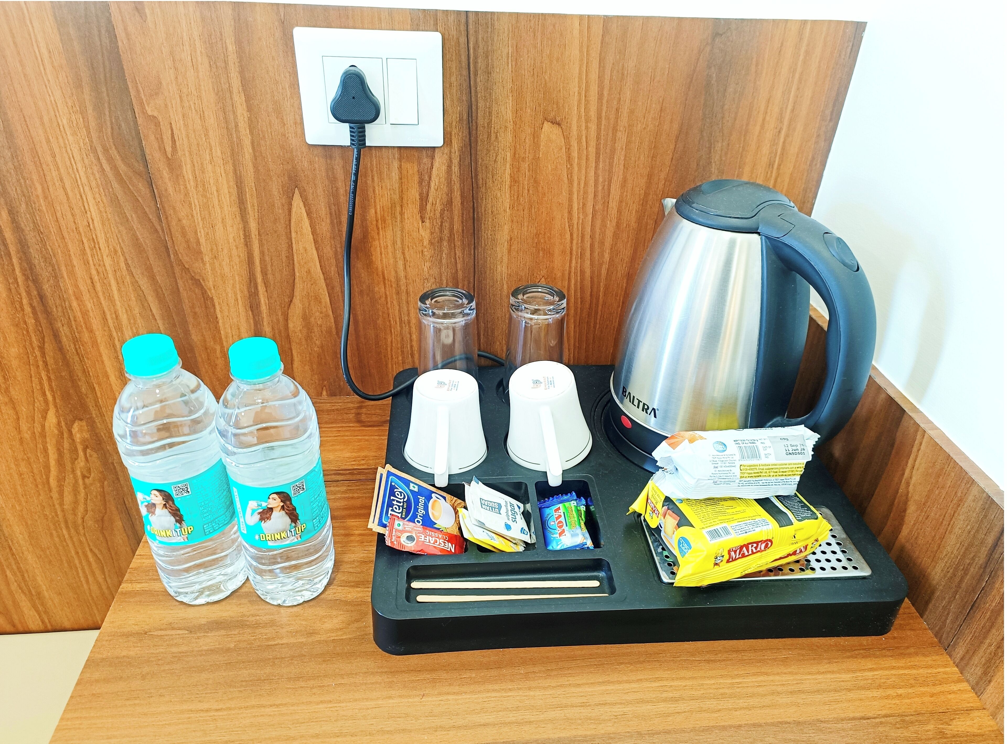 Uma cozinha compacta com chaleira, duas garrafas de água, cafeteira, bule, caneca, caixa de saquinhos de chá, sachê de açúcar e sachê de chá.