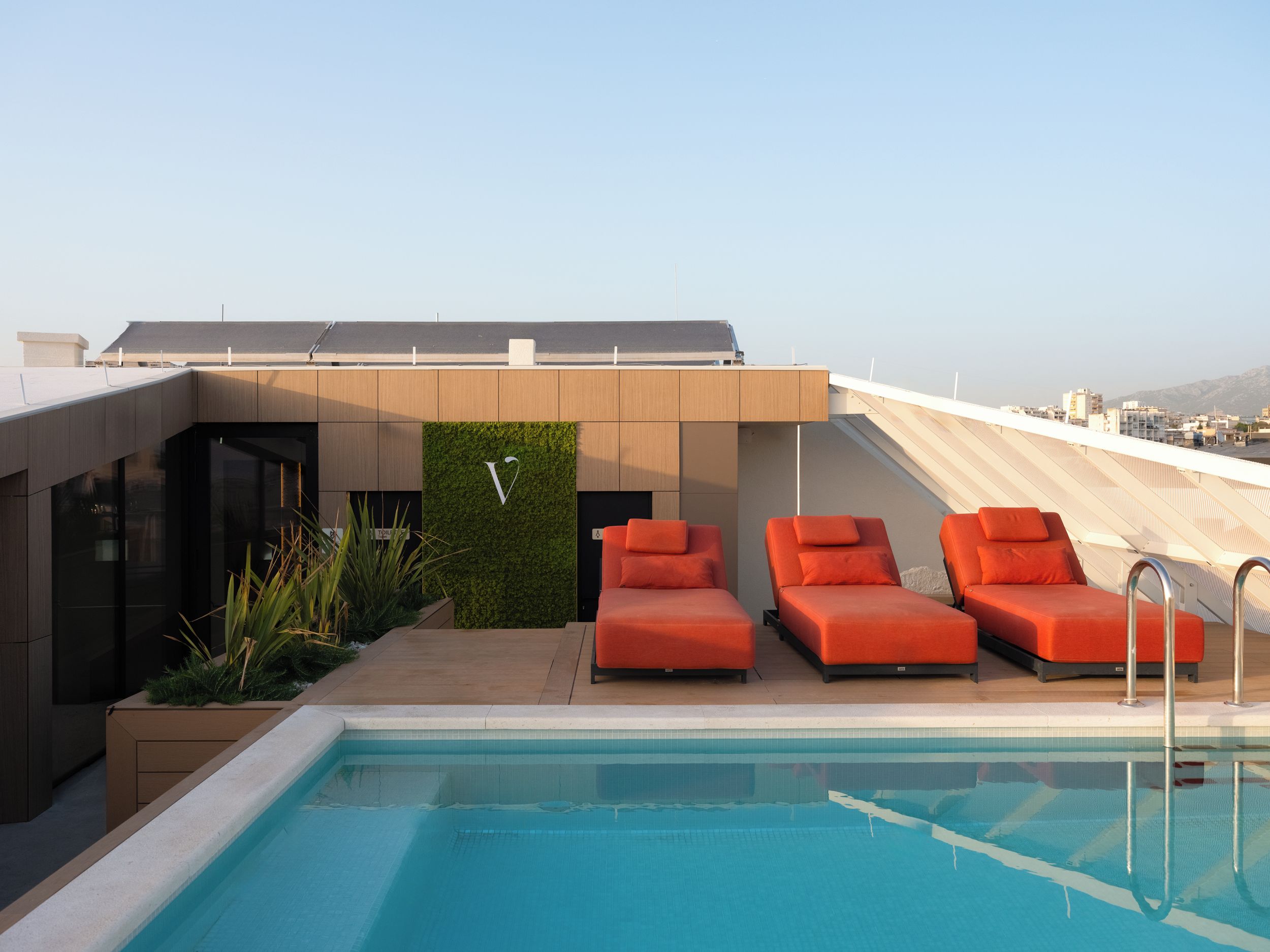Área de piscina no terraço, com três cadeiras de sol laranja e uma parede verde com a letra 'V'.