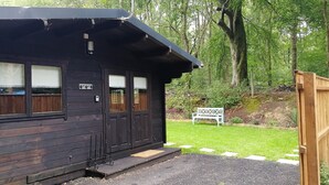 Uma cabana de madeira com exterior escuro, um pequeno alpendre e um banco branco no quintal.