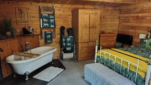 Interior de cabana de madeira com banheira, cama com cabeceira amarela, guarda-roupa de madeira e pequena mesa com luminária.