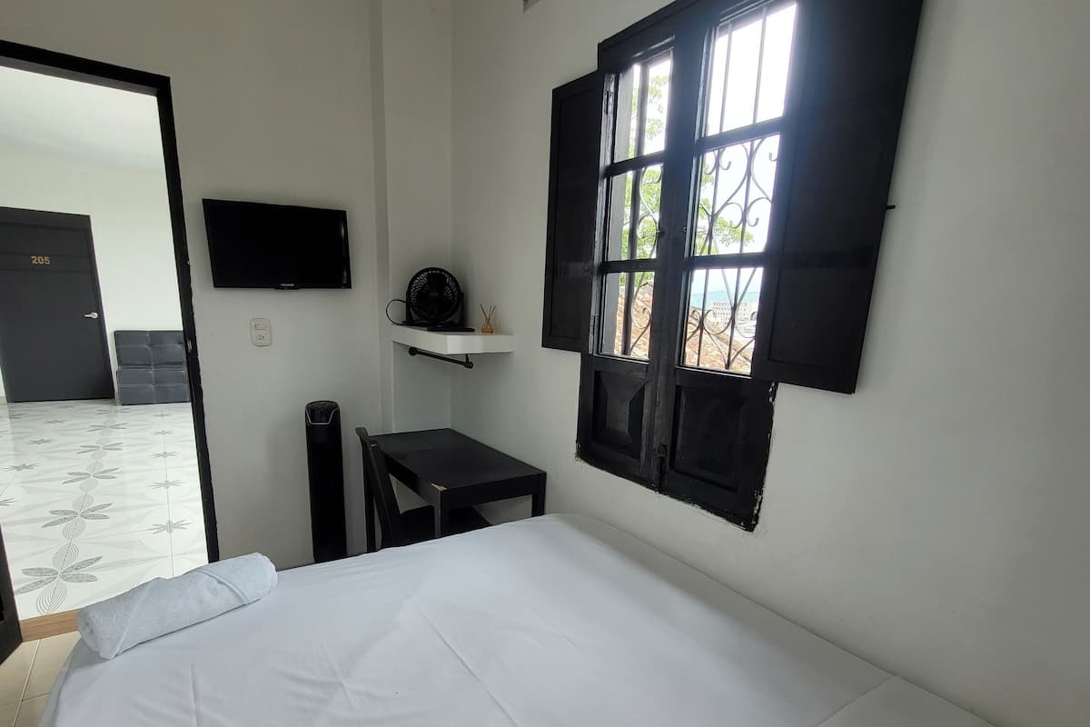 Quarto de hotel com cama, escrivaninha, televisão e janela com portadas.