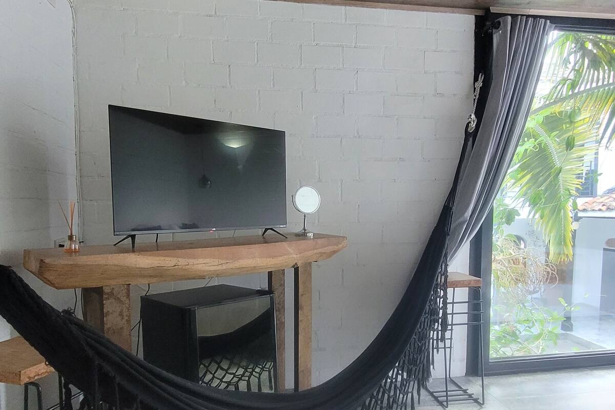 Uma rede, uma televisão e uma pequena mesa em um quarto com parede de tijolos brancos e teto de madeira.