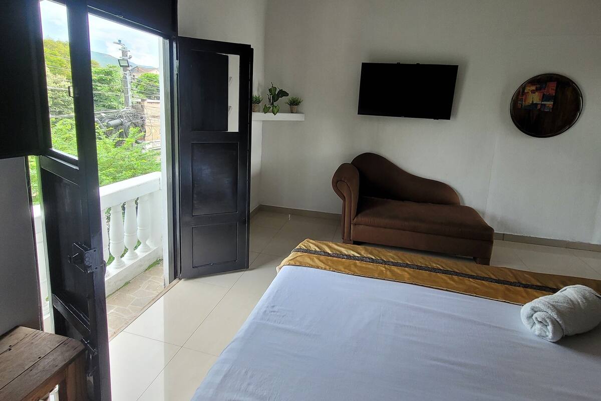 Quarto de hotel com cama, um sofá marrom, televisão fixada na parede e janela com vista para o verde.