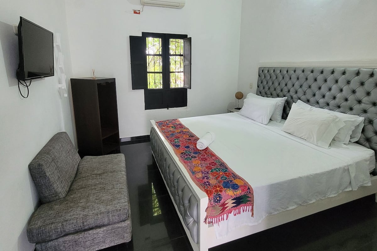Quarto com cama, poltrona estofada cinza, televisão e janela com persianas.