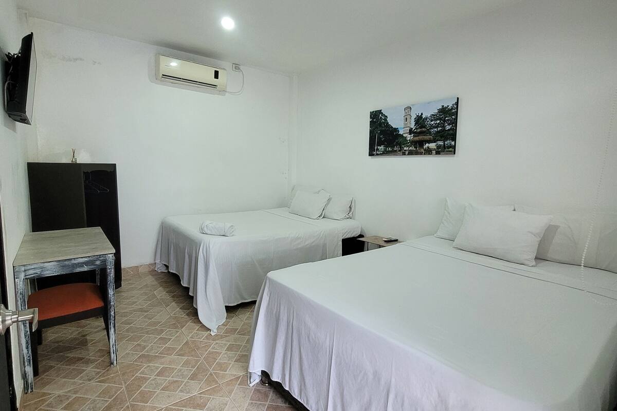 Quarto de hotel com duas camas de solteiro, uma mesa pequena, ar condicionado e televisão presa à parede.