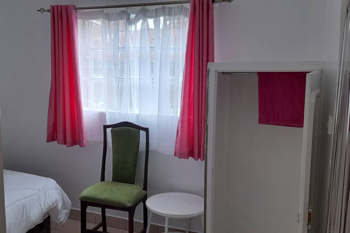 Um quarto pequeno com uma cama, uma cadeira, uma mesa e uma janela com cortinas.