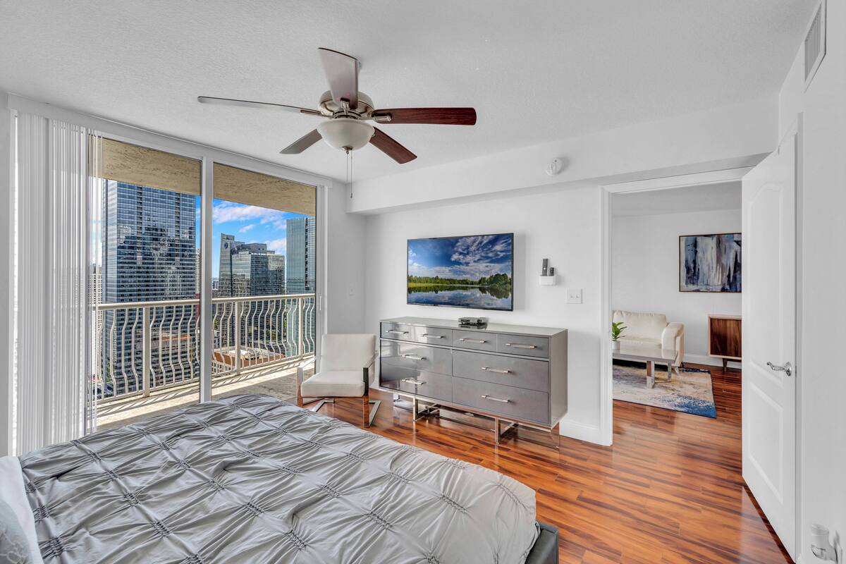 Quarto moderno com cama grande, ventoinha no teto, varanda com vista para a cidade e televisão.