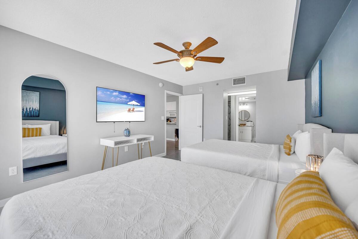 Quarto de hotel com duas camas, um ventilador de teto, uma televisão, uma escrivaninha e a porta do banheiro.