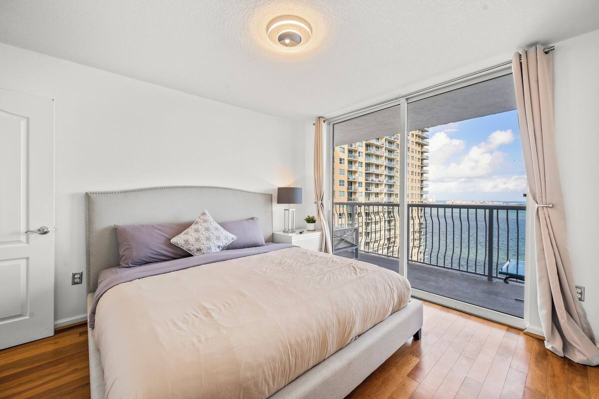 Quarto com cama grande, mesa de cabeceira, luminária e vista para um edifício alto através de uma porta de vidro deslizante.