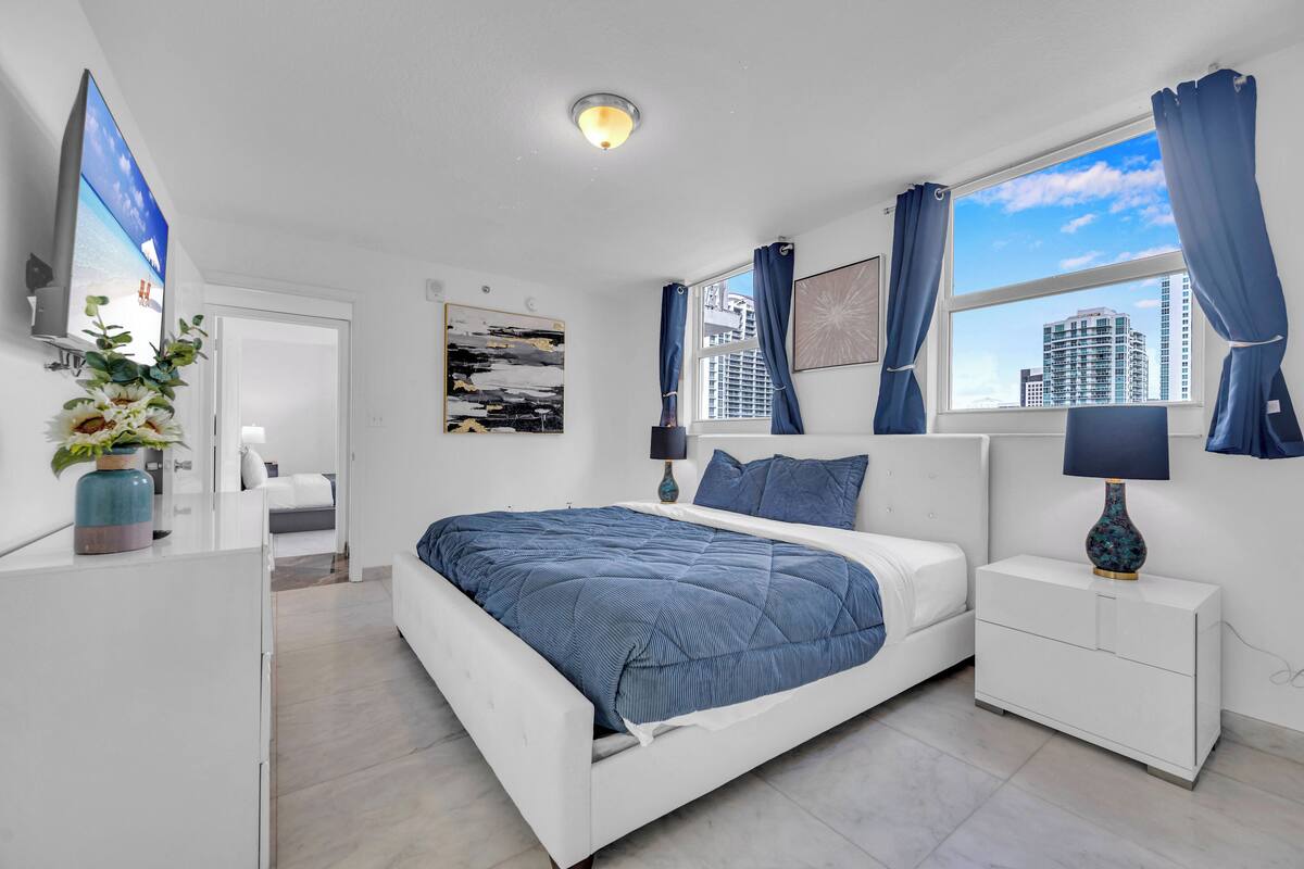 Quarto moderno com uma cama grande, mesa de cabeceira com luminária, cômoda, televisão na parede e uma janela com cortinas.