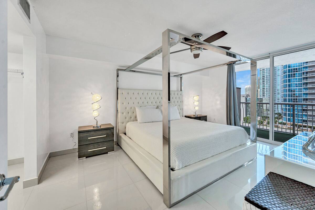 Quarto moderno com cama de dossel em vidro, cabeceira estofada, abajures e vista para a cidade.