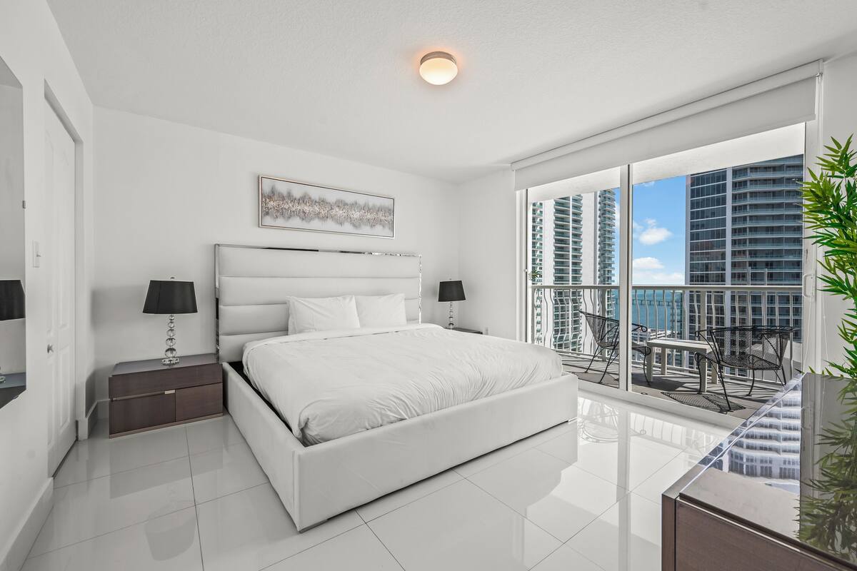 Quarto moderno com uma cama grande, duas mesas de cabeceira, uma mesinha de apoio, uma janela com vista para edifícios altos e uma luminária no teto.