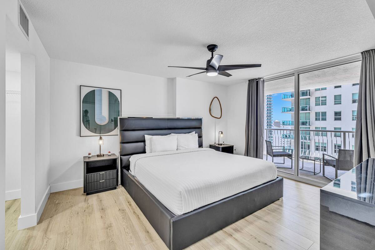 Quarto de hotel moderno com uma cama grande, um ventilador de teto, uma janela ampla com vista para edifícios e uma pequena mesa de trabalho com uma luminária.