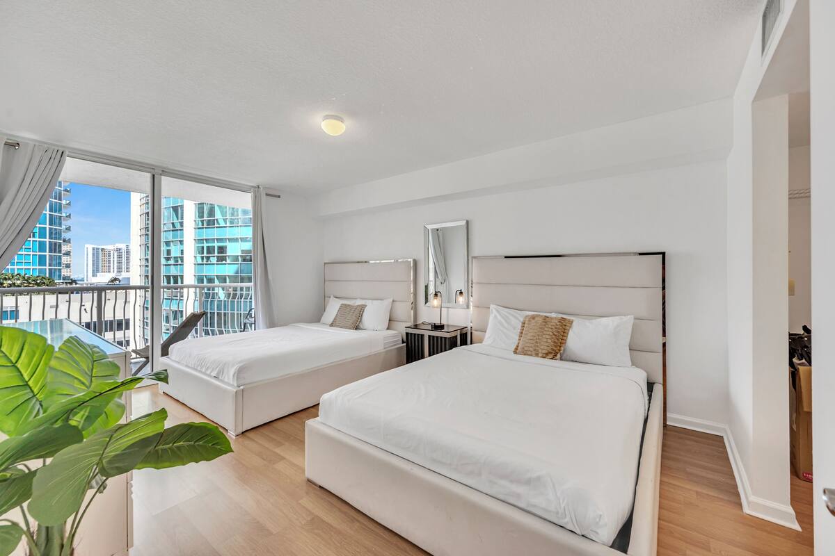 Quarto de hotel moderno com duas camas, varanda e vista para a cidade, além de uma planta grande.