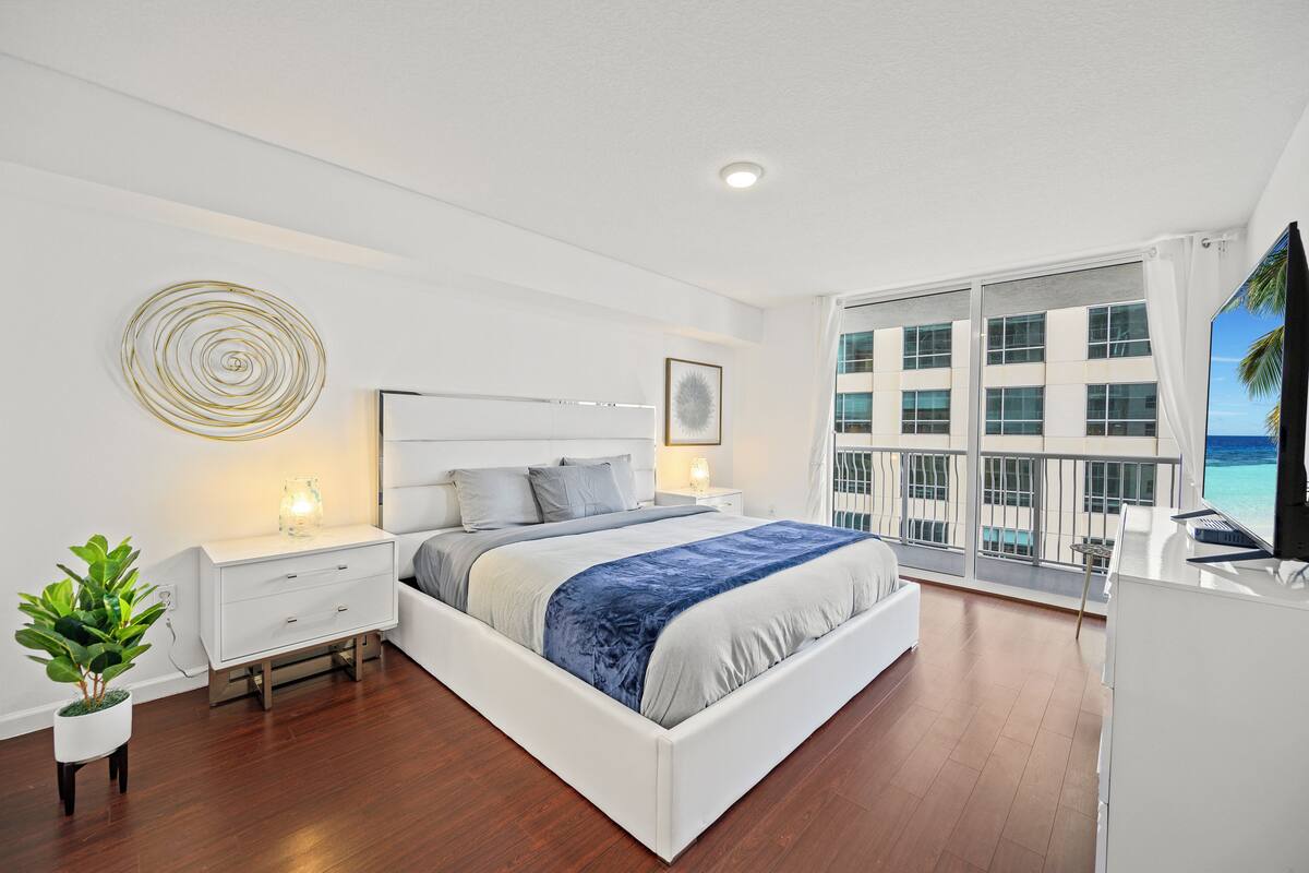 Quarto moderno com uma cama grande, uma mesa de cabeceira, uma planta em vaso e vista para o oceano através de amplas janelas.