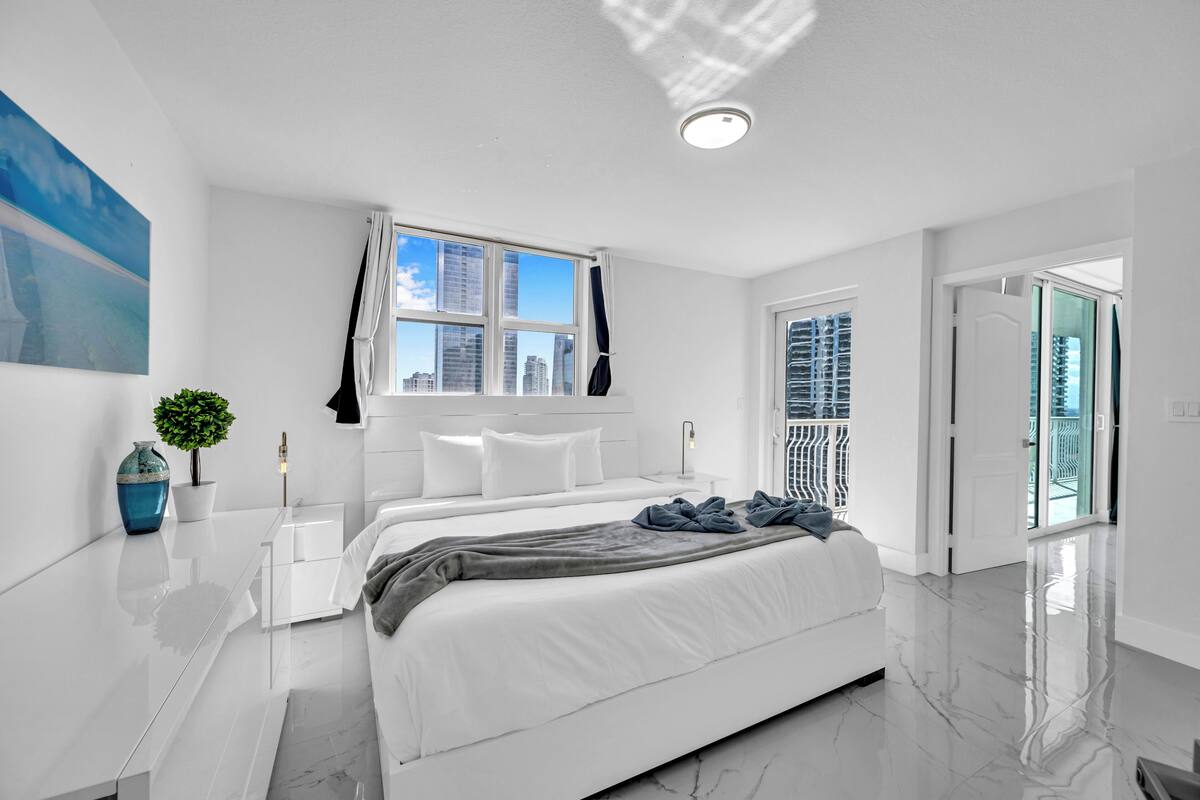 Quarto moderno com uma cama grande, uma mesinha de cabeceira, um vaso com planta e vista para a cidade pela janela.