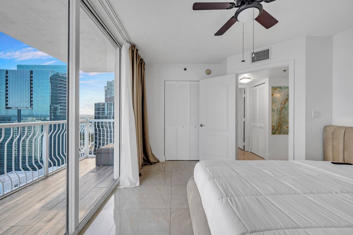 Quarto de hotel com varanda, cama, ventilador de teto e vista para edifícios da cidade.