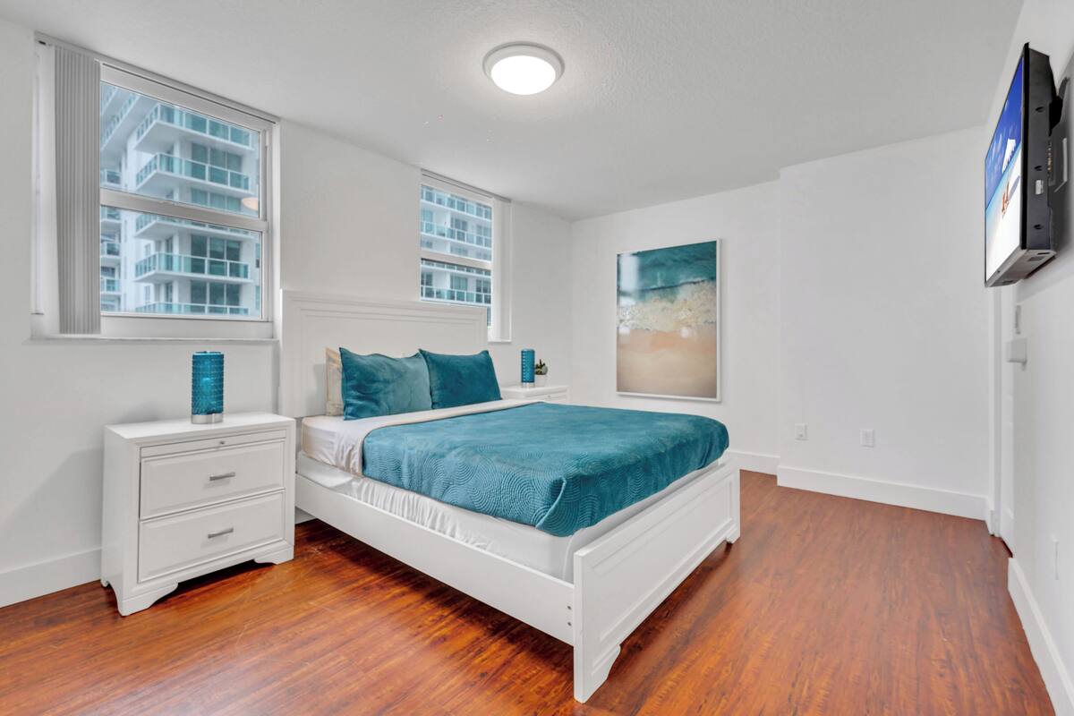 Quarto com cama branca, mesa de cabeceira, televisão fixada na parede e duas janelas com vista para um edifício.