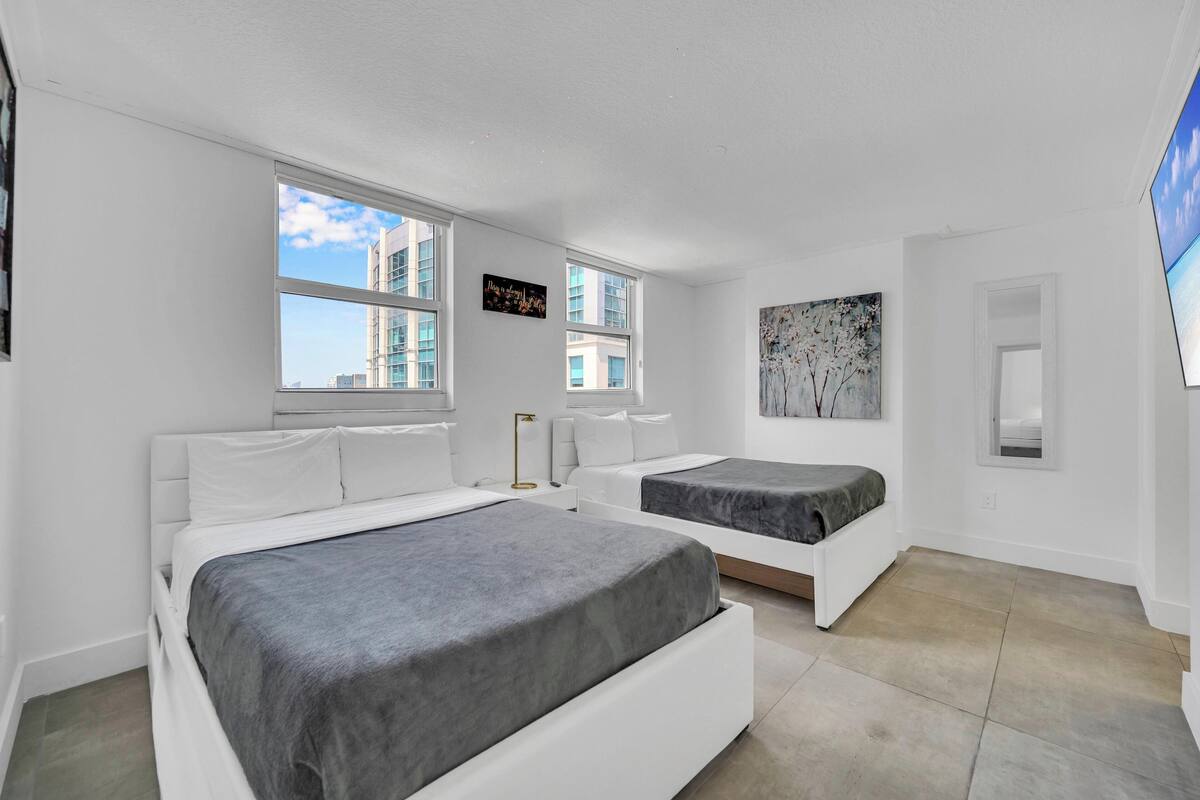Quarto moderno com duas camas, janelas amplas, um quadro e uma televisão.