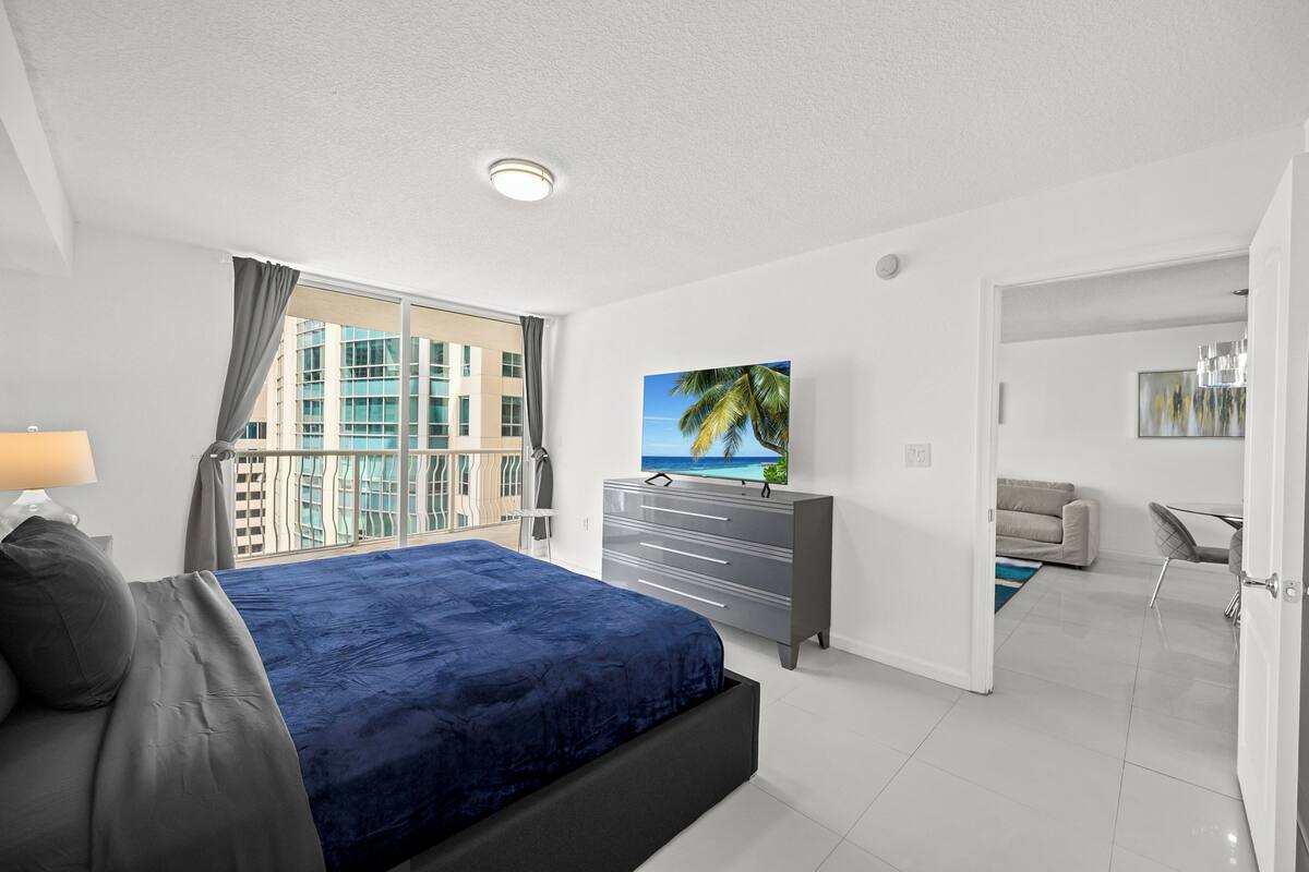 Quarto moderno com uma cama grande, cômoda, TV e varanda com vista para a cidade.