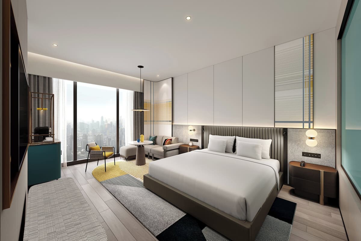 Quarto de hotel moderno com uma cama grande, área de estar com sofá e poltronas, uma mesa pequena e vista para a cidade através de grandes janelas.