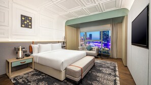 Quarto de hotel com uma cama grande, uma mesa de cabeceira com abajur, uma cadeira, uma pequena mesa e vista para a paisagem urbana à noite.
