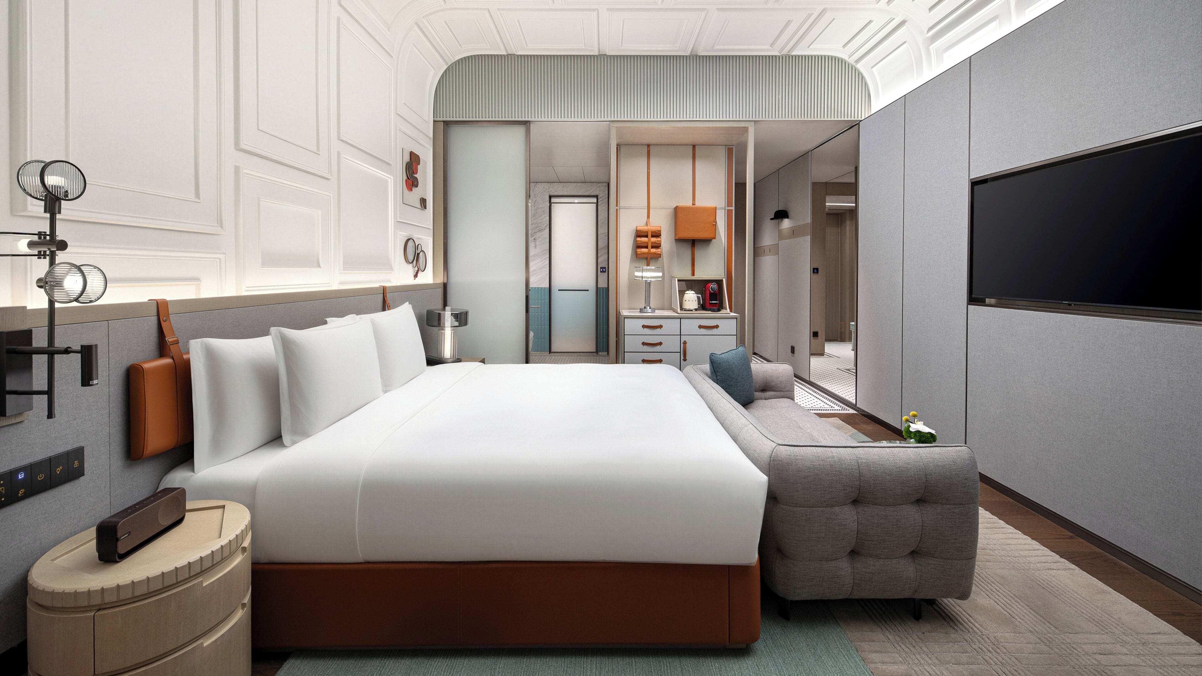 Quarto de hotel moderno com uma cama grande, uma mesa com abajur, um sofá e uma televisão.