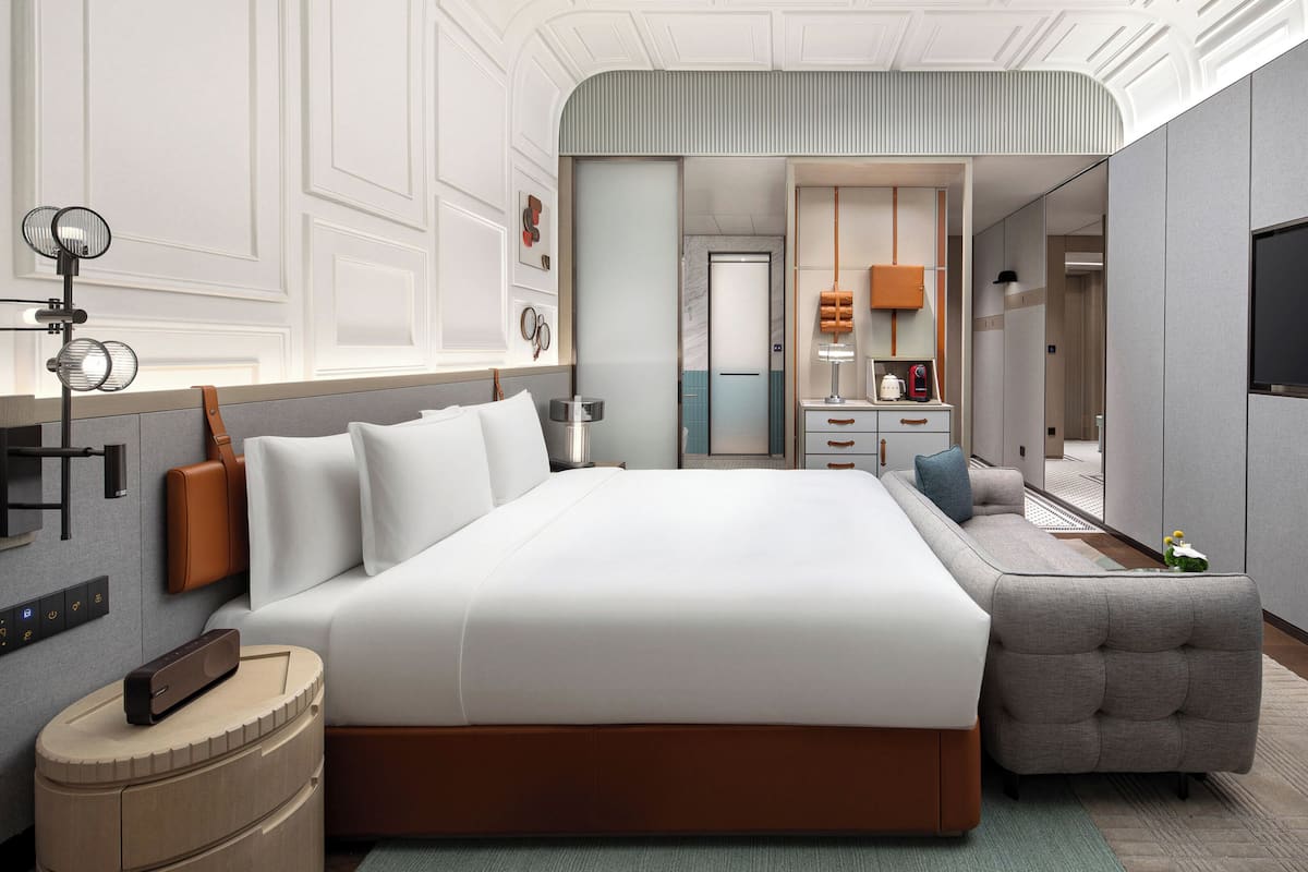 Quarto de hotel moderno com uma cama grande, uma mesa com abajur, um sofá e uma televisão.