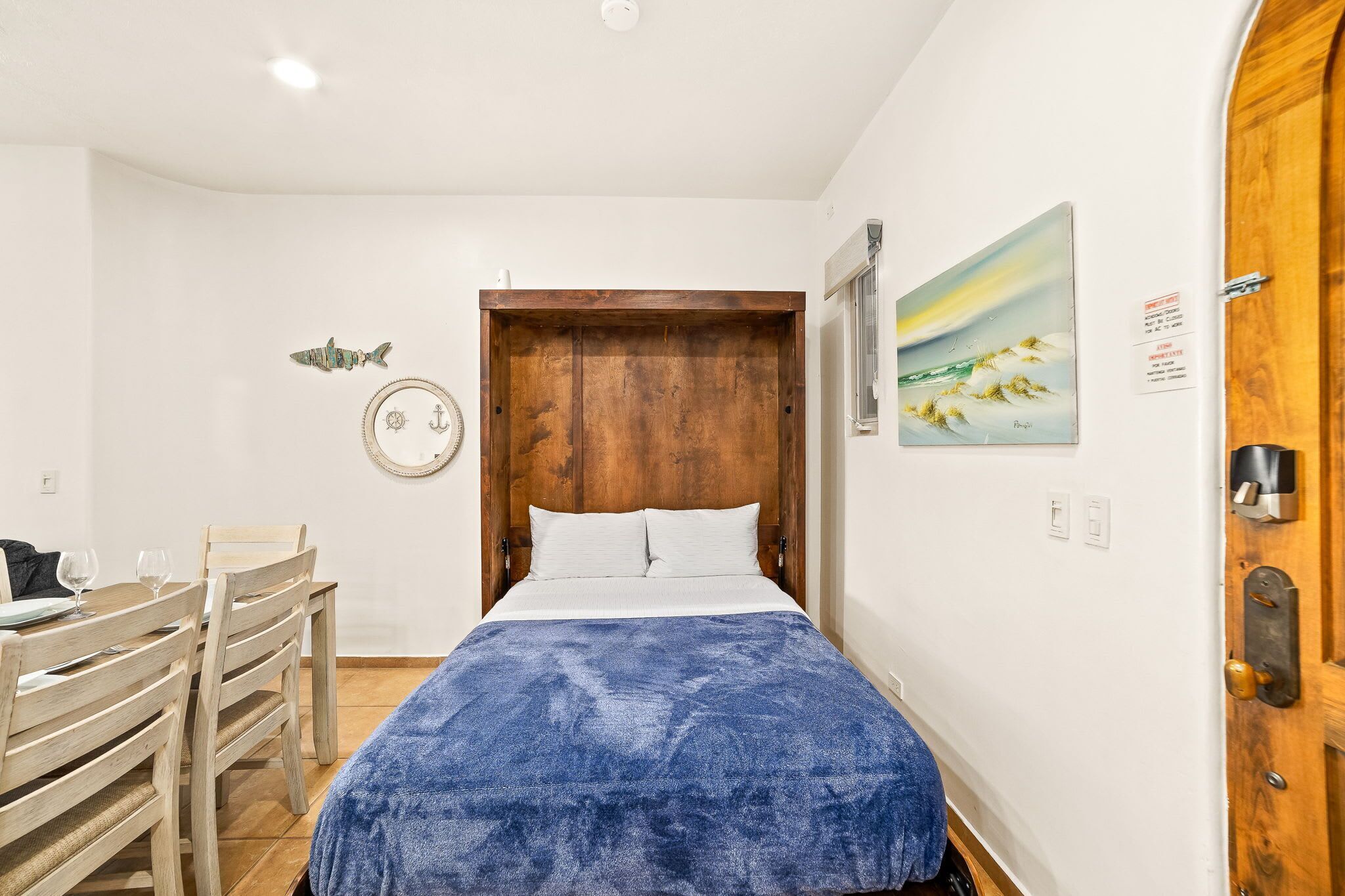 Quarto com cabeceira de madeira, cama com cobertor azul, mesa de jantar de madeira com cadeiras e um quadro pendurado na parede.