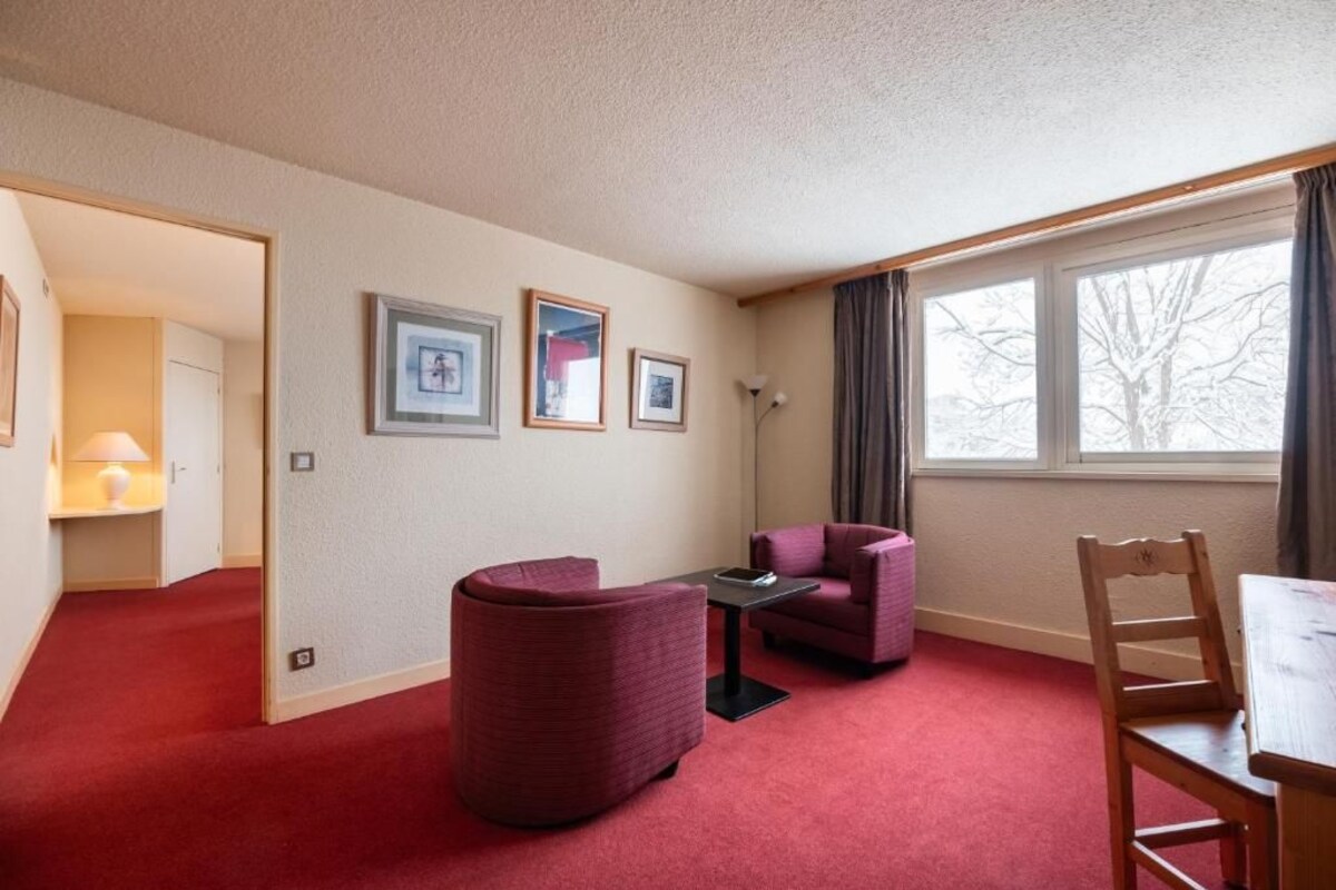 Quarto de hotel com tapete vermelho, sofá, cadeira, mesinha e janela com cortinas.