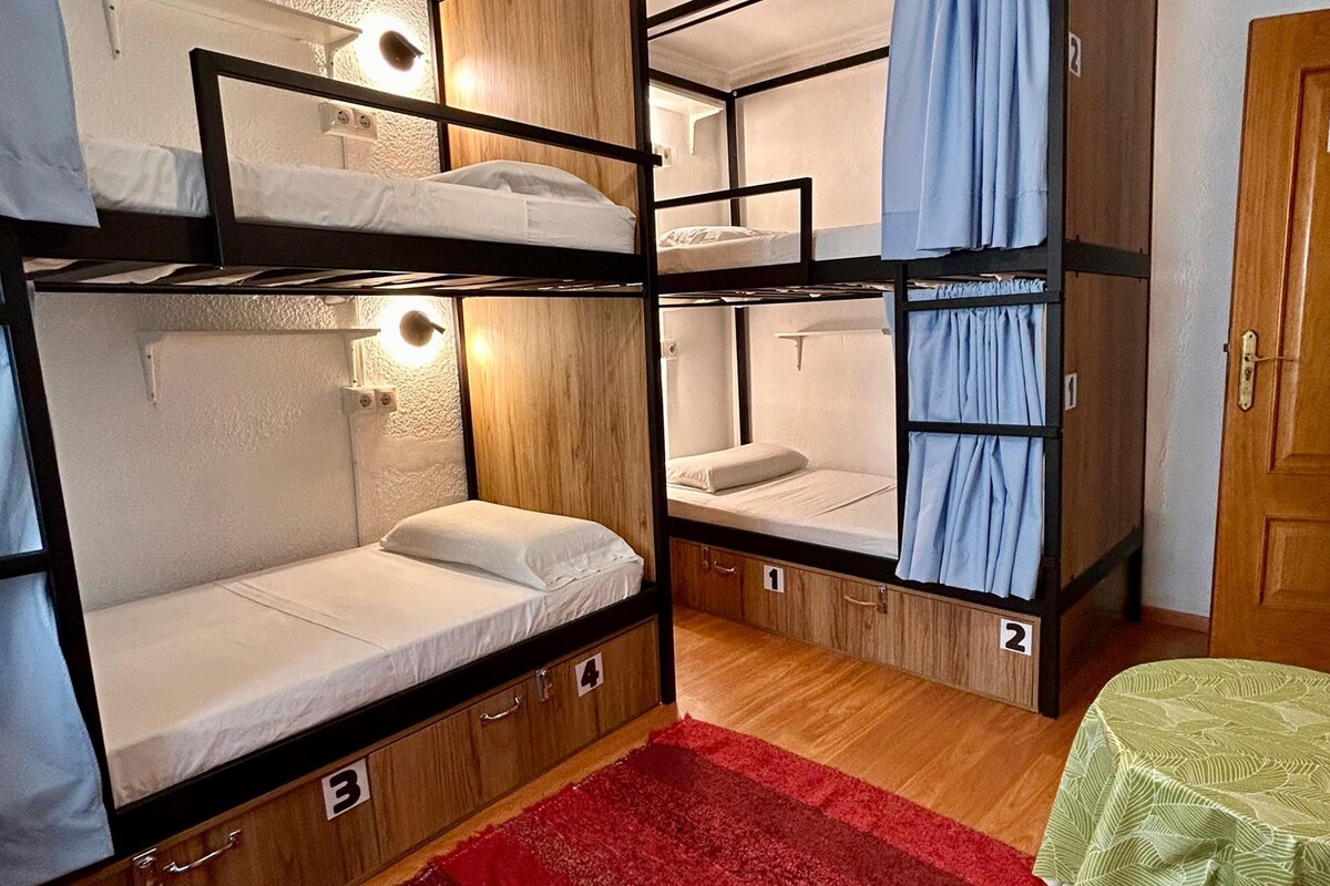 Quarto com beliches, tapete vermelho e uma pequena mesa com toalha verde.