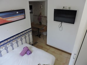 Quarto de hotel com uma cama, televisão fixada na parede, um quadro na parede e um pequeno espaço de refeições com uma mesa e cadeiras.