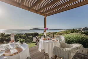 Área de refeições ao ar livre coberta, com mesas preparadas para o café da manhã e vista para a costa.