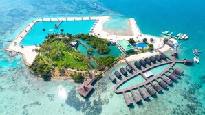 Vista aérea de uma ilha resort com águas turquesa cristalinas, piscina central e bangalôs sobre a água.