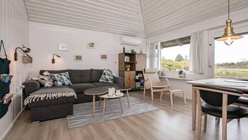 Uma sala de estar acolhedora com um sofá cinza escuro, uma mesa redonda pequena e uma mesa de jantar de madeira com cadeiras. Há uma estante cheia de livros e objetos decorativos, e uma janela com vista para o verde lá fora.