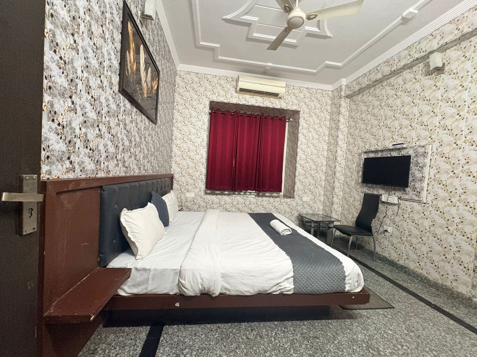 Quarto de hotel com cama, escrivaninha, cadeira, televisão e janela com cortinas vermelhas.