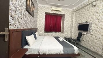 Quarto de hotel com cama, escrivaninha, cadeira, televisão e janela com cortinas vermelhas.