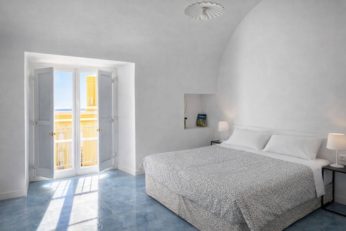 Um quarto com cama, duas mesas de cabeceira, uma mesinha de apoio, uma porta que dá para uma varanda e um lustre no teto.
