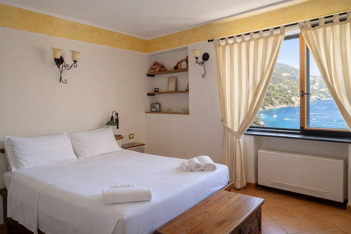 Quarto com cama, mesas de cabeceira, janela com cortinas e vista para o mar.