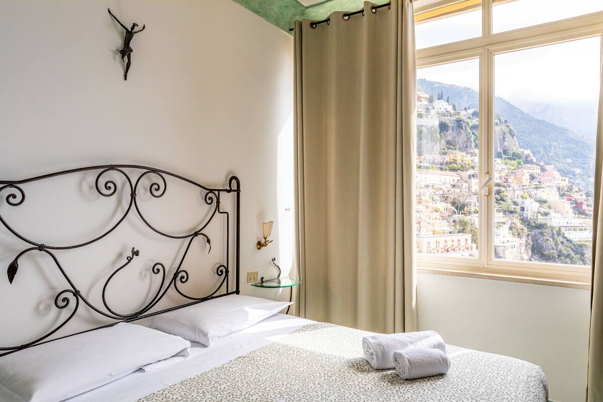 Uma cama com cabeceira de metal, uma mesa de cabeceira com abajur e vista para uma cidade no topo de uma colina através da janela.