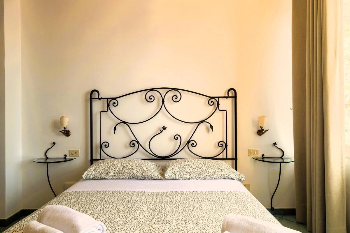 Uma cama com cabeceira metálica decorativa, duas mesas de cabeceira com luminárias e um edredom estampado.