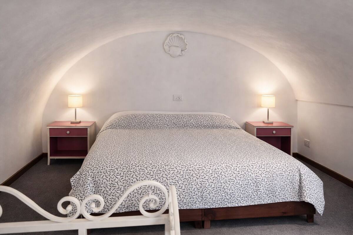Uma cama de solteiro num quarto com teto abobadado, duas mesas de cabeceira com abajures e uma concha decorativa na parede.