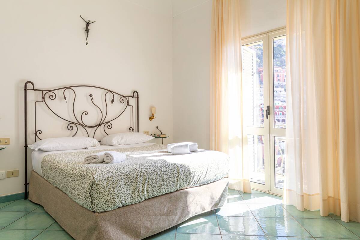Uma cama com cabeceira de metal, uma mesa de cabeceira com luminária e vista através de uma porta de vidro.