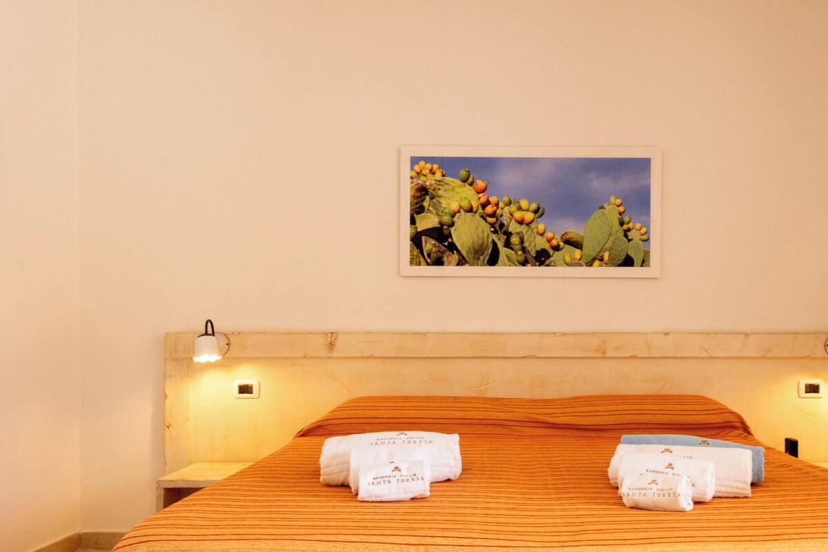 Quarto de hotel com cama, duas luminárias de cabeceira, um quadro com cactos e duas toalhas sobre a cama.